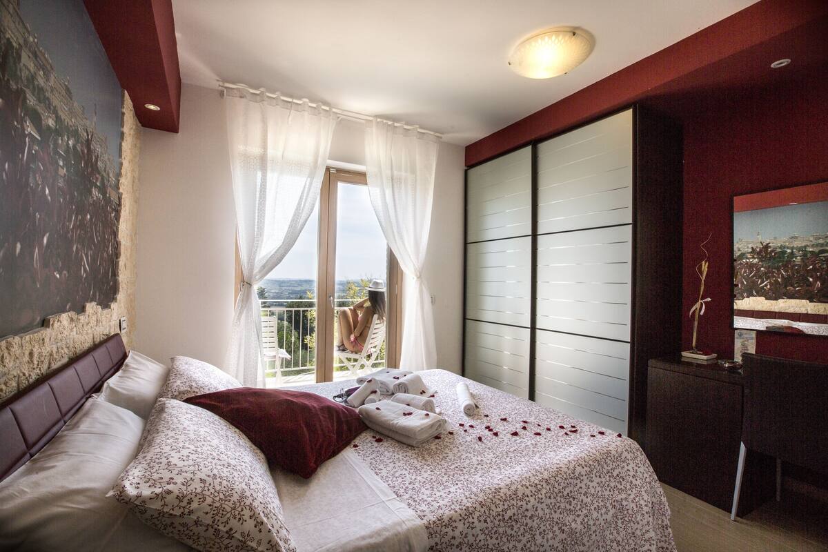 Quarto com cama, escrivaninha, cadeira, janela com cortinas e vista para o exterior.
