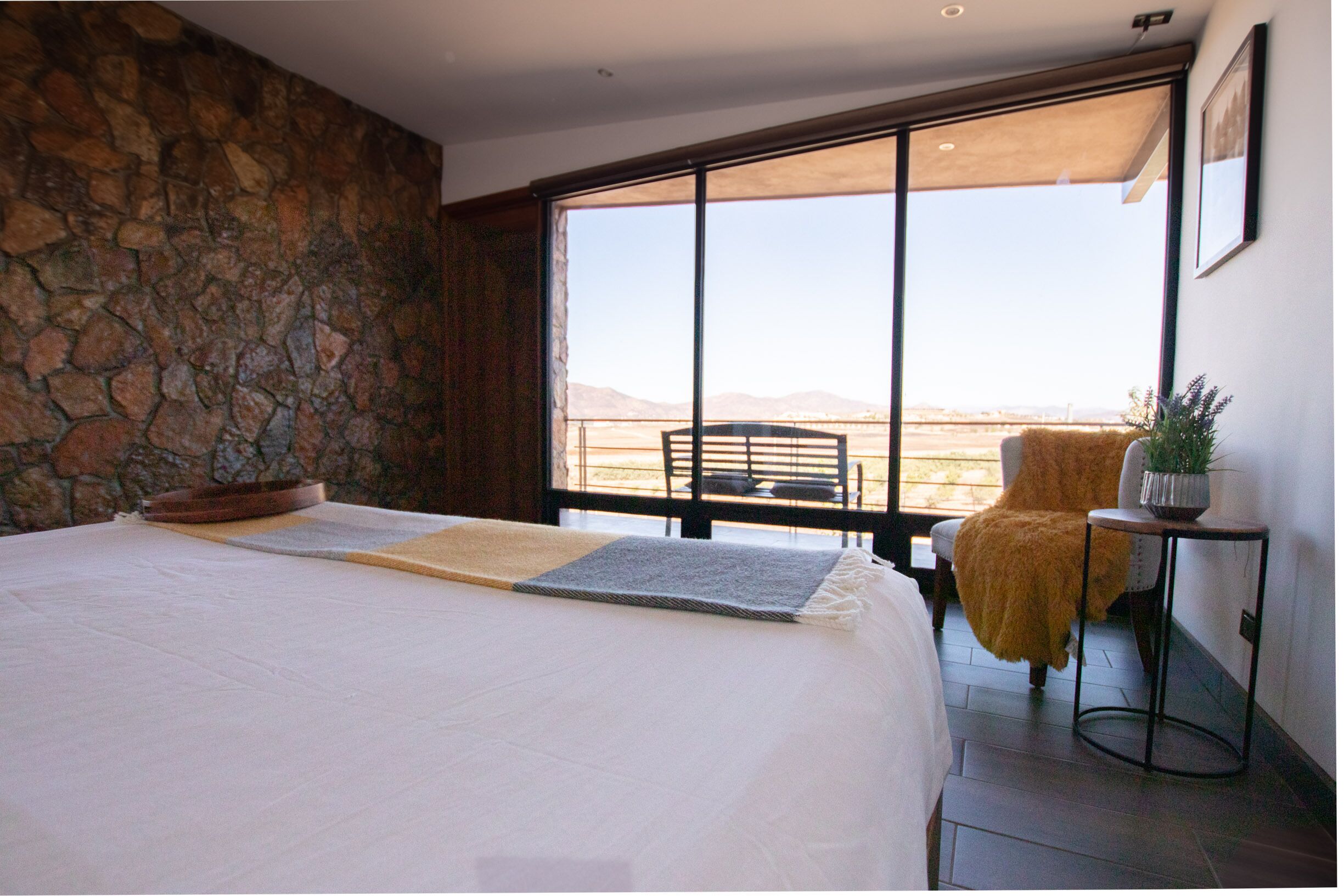 Quarto com uma cama grande, parede de pedra, vista através de uma janela ampla e uma mesinha com uma planta.