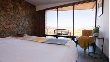 Quarto com uma cama grande, parede de pedra, vista através de uma janela ampla e uma mesinha com uma planta.