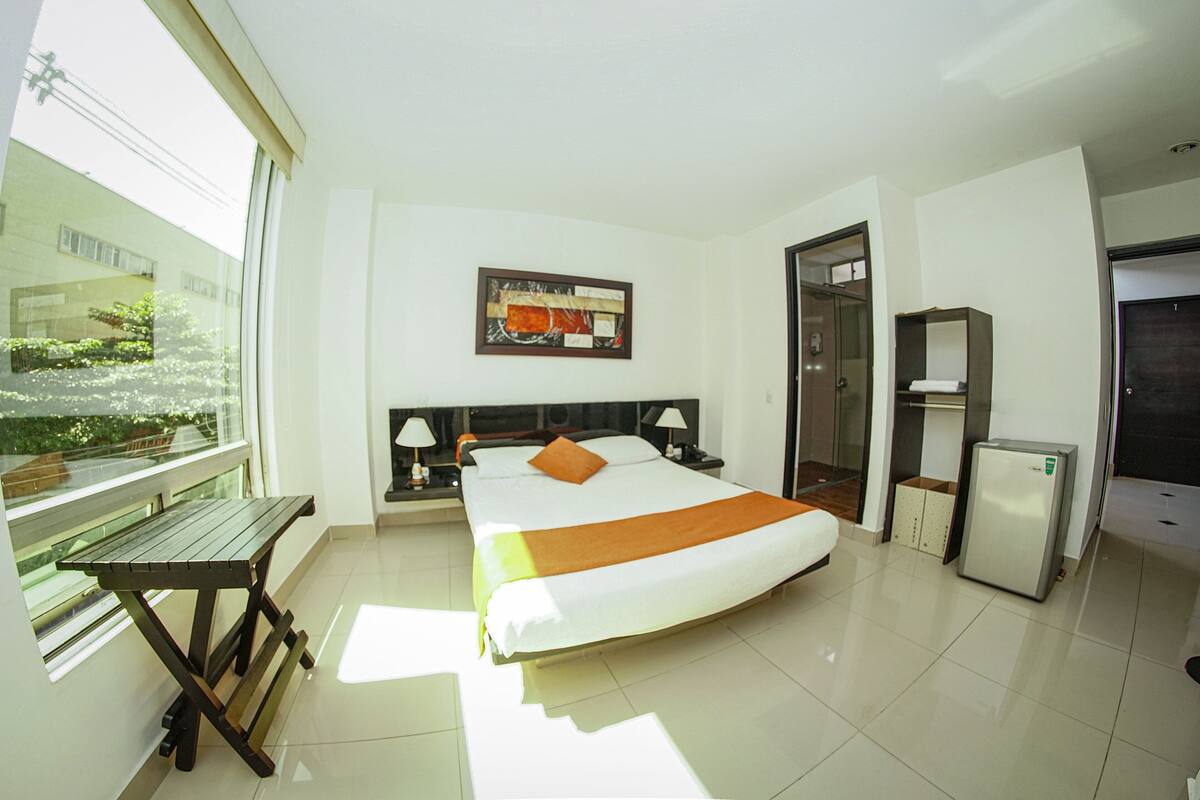 Quarto de hotel com cama, mesa de cabeceira, abajur e um pequeno frigorífico.