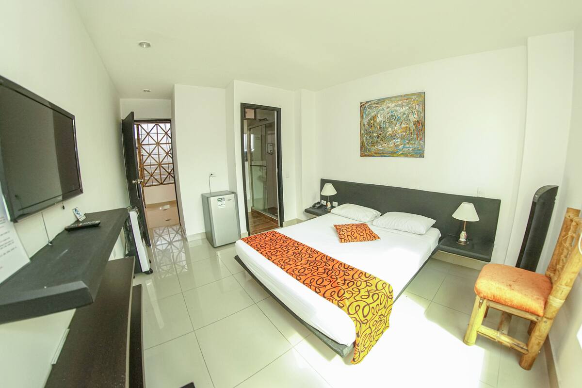 Quarto de hotel com cama, mesa de cabeceira, abajur, cadeira, escrivaninha, televisão e porta para uma varanda.
