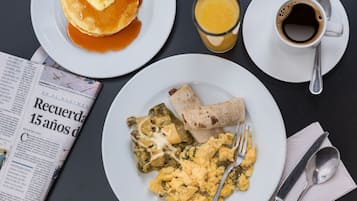 Prato de café da manhã com panquecas, ovos mexidos e um wrap, uma xícara de café e um copo de suco de laranja sobre uma mesa com um jornal.