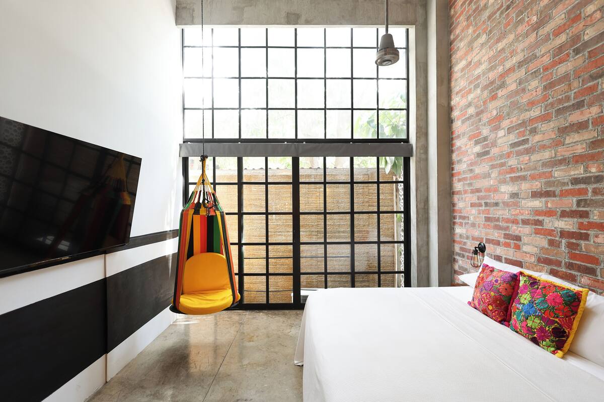 Quarto de hotel moderno com uma televisão grande, uma cama com almofadas coloridas, uma cadeira suspensa e uma parede de tijolos.