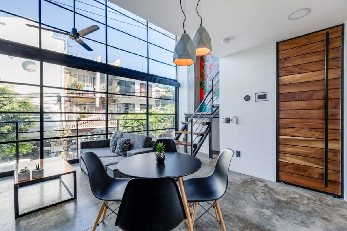 Quarto moderno com uma janela grande, uma mesa redonda com cadeiras, um ventilador de teto e uma escada com corrimão colorido.