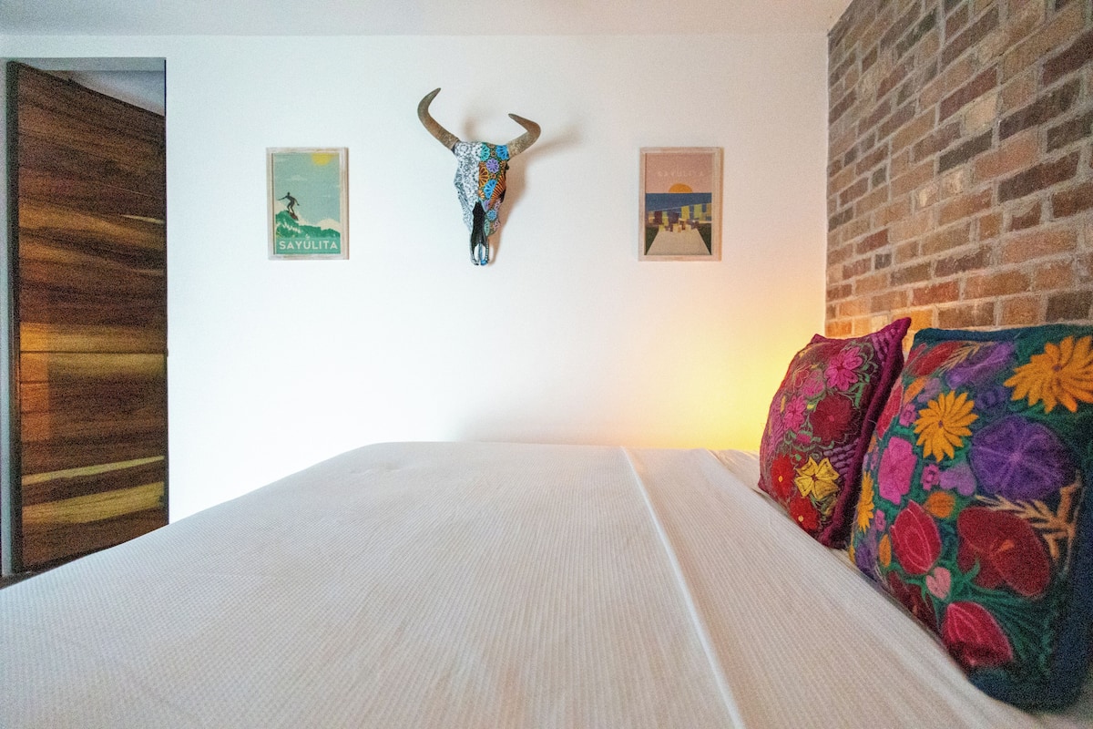 Uma cama com lençóis brancos, uma porta de madeira, uma parede de tijolos e duas molduras com quadros na parede.