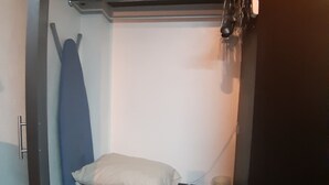 Quarto compacto com uma cama, um pequeno armário com objetos e uma porta.
