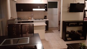 Cozinha moderna com fogão a gás, micro-ondas e frigorífico. Possui área de jantar com mesa e cadeiras. Uma televisão de ecrã plano está fixada na parede.
