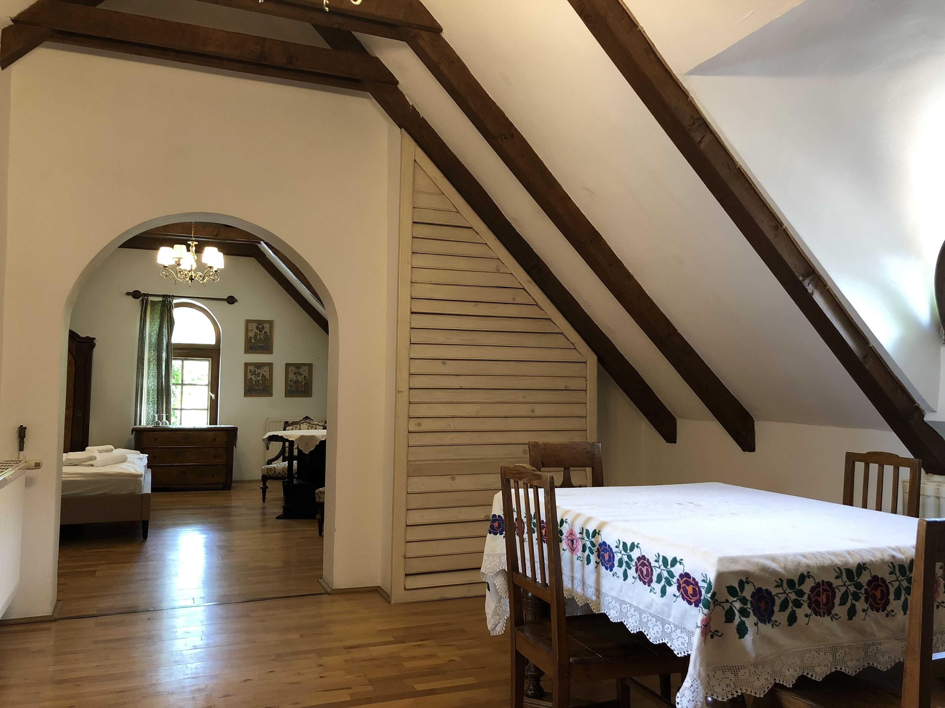 Quarto com teto inclinado, vigas de madeira, uma mesa de jantar com toalha florida, uma cadeira de madeira e vista através de uma porta arqueada para outro quarto com uma cama e uma janela.
