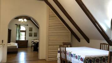 Quarto com teto inclinado, vigas de madeira, uma mesa de jantar com toalha florida, uma cadeira de madeira e vista através de uma porta arqueada para outro quarto com uma cama e uma janela.