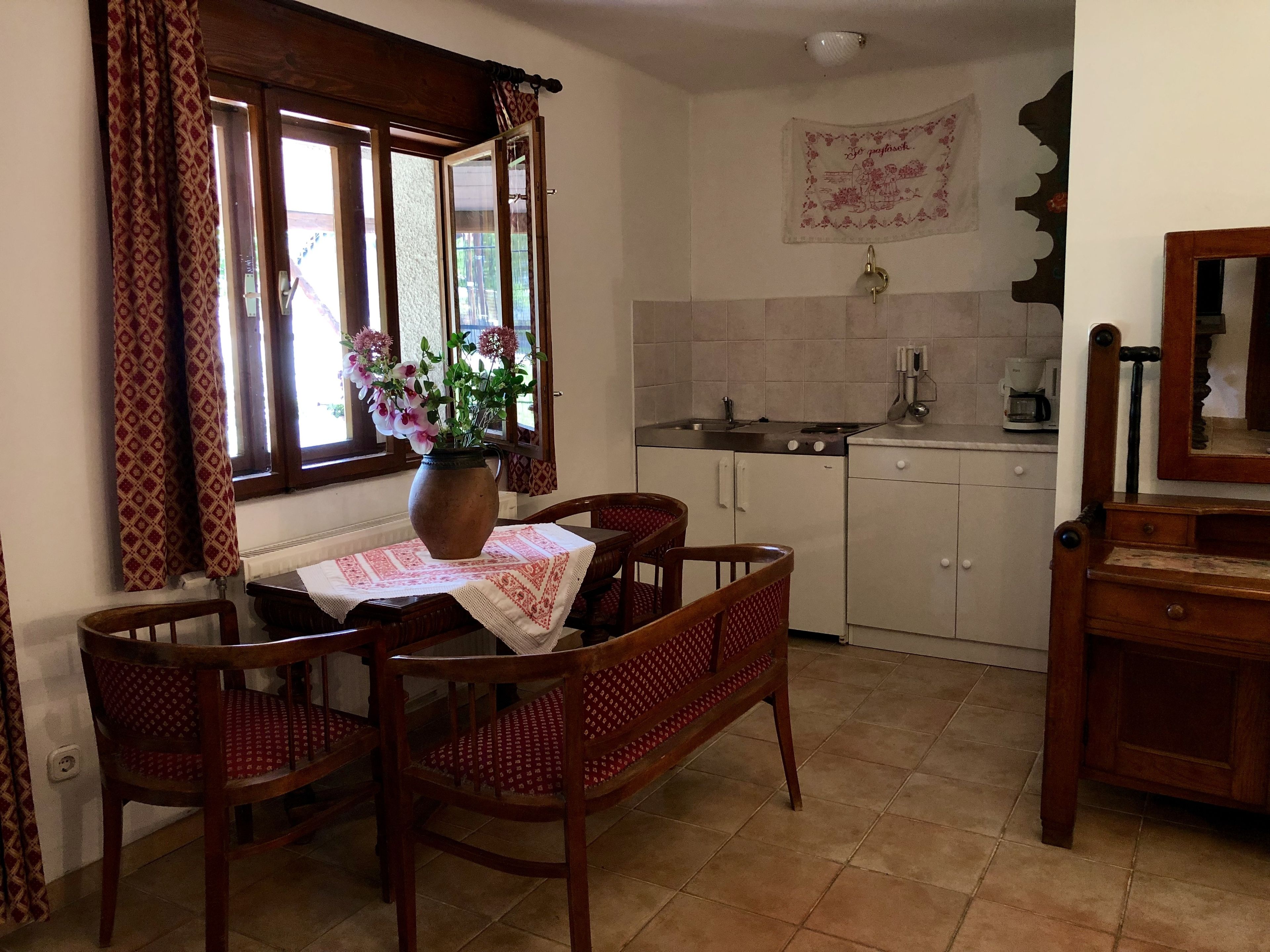 Uma cozinha com área de jantar, destacando uma mesa com toalha estampada, cadeiras de madeira, um vaso com flores e uma janela com cortinas.