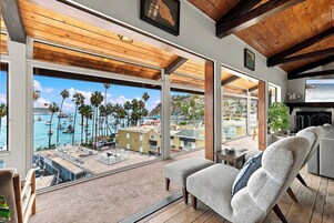Quarto com amplas janelas para uma marina e palmeiras, vigas de madeira no teto e lareira.