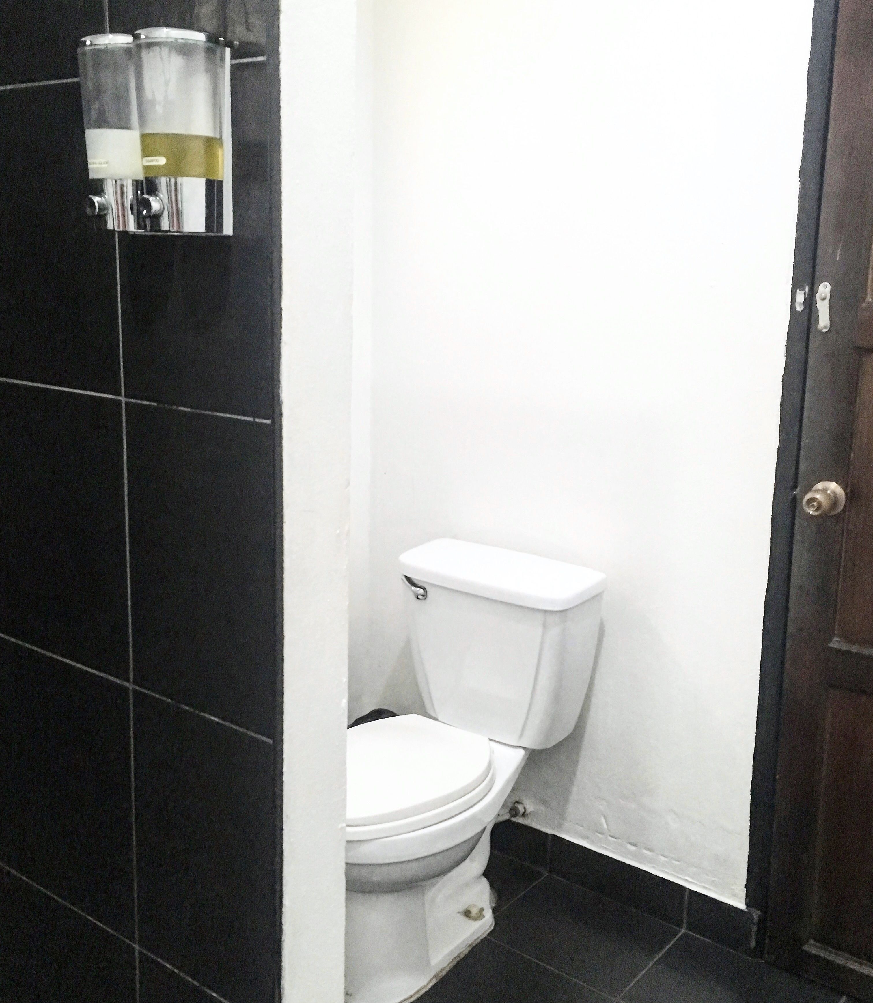 Uma sanita branca com depósito cromado, parede revestida a azulejos pretos e porta com puxador prateado.