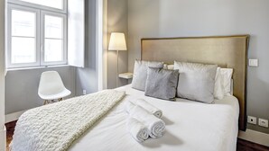 Quarto de hotel com uma cama grande, uma cadeira branca, mesa de cabeceira com abajur e janela com cortinas.