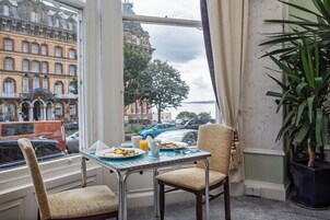 Mesa de café da manhã com pratos, jarra e xícara, perto de uma janela com vista para um horizonte urbano com edifícios históricos e carros estacionados.