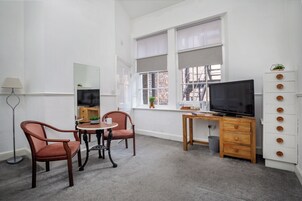 Quarto com televisão, duas cadeiras, uma mesinha, luminária, janela com persianas e cómoda com gavetas.