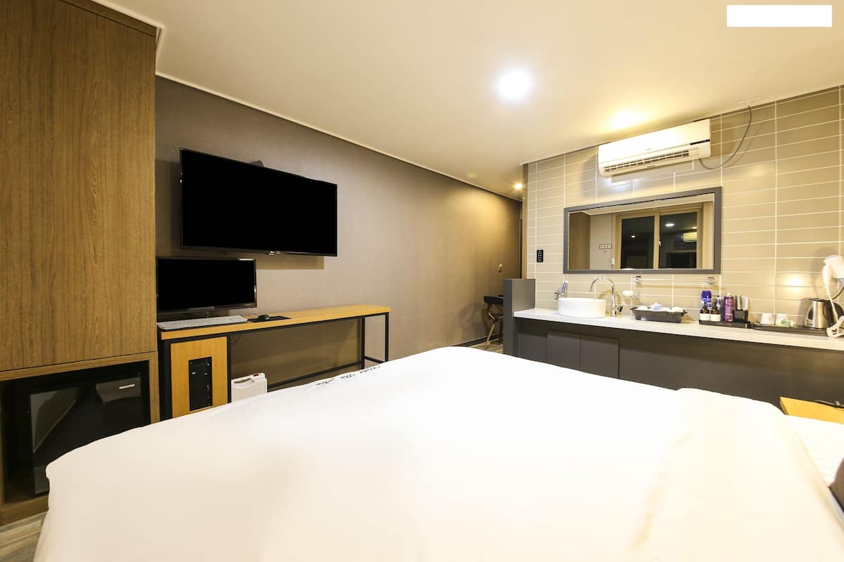 Quarto de hotel com cama, televisão, escrivaninha, mini-geladeira, pia e espelho.