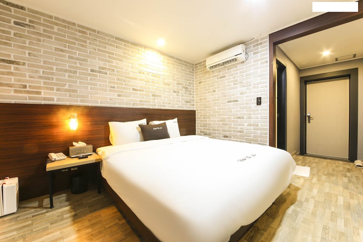 Quarto de hotel com parede em padrão de tijolos, cabeceira de madeira, cama branca, mesa de cabeceira com telefone e luminária, e unidade de ar condicionado.