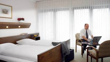 Quarto de hotel com uma cama, uma escrivaninha e uma cadeira. Há uma janela com cortinas leves, uma estante com livros e um vaso de flores, e um quadro na parede.