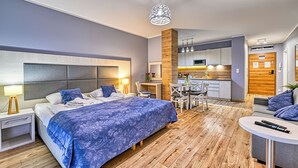 Quarto de hotel moderno com uma cama grande, uma escrivaninha, uma cadeira, pequena cozinha e área de estar com mesa e cadeiras.