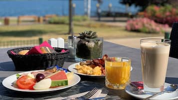 Uma mesa posta com itens de café da manhã, incluindo torradas, ovos, bacon e frutas, com vista para um lago ao fundo.