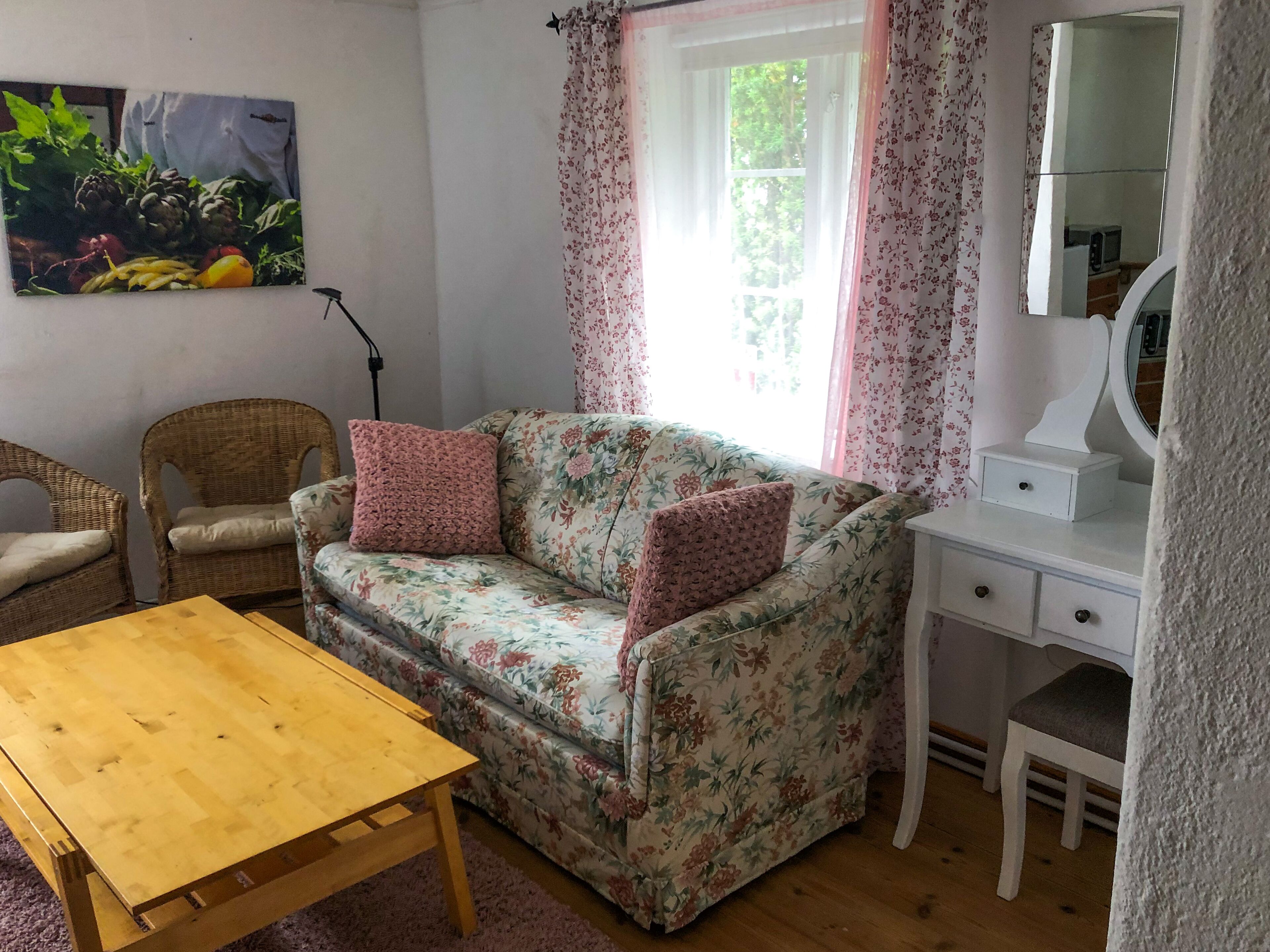 Uma sala de estar acolhedora com um sofá floral, duas almofadas combinando, uma mesa de centro de madeira, uma escrivaninha branca com espelho e uma cadeira de vime.