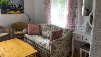 Uma sala de estar acolhedora com um sofá floral, duas almofadas combinando, uma mesa de centro de madeira, uma escrivaninha branca com espelho e uma cadeira de vime.