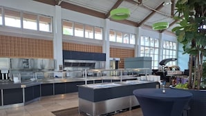 Um café espaçoso com cozinha moderna, área de estar com mesas e cadeiras, e amplas janelas que proporcionam iluminação natural.