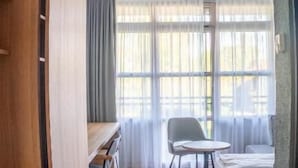 Quarto de hotel com guarda-roupa de madeira, uma pequena escrivaninha com cadeira, uma cama e uma janela grande com cortinas.