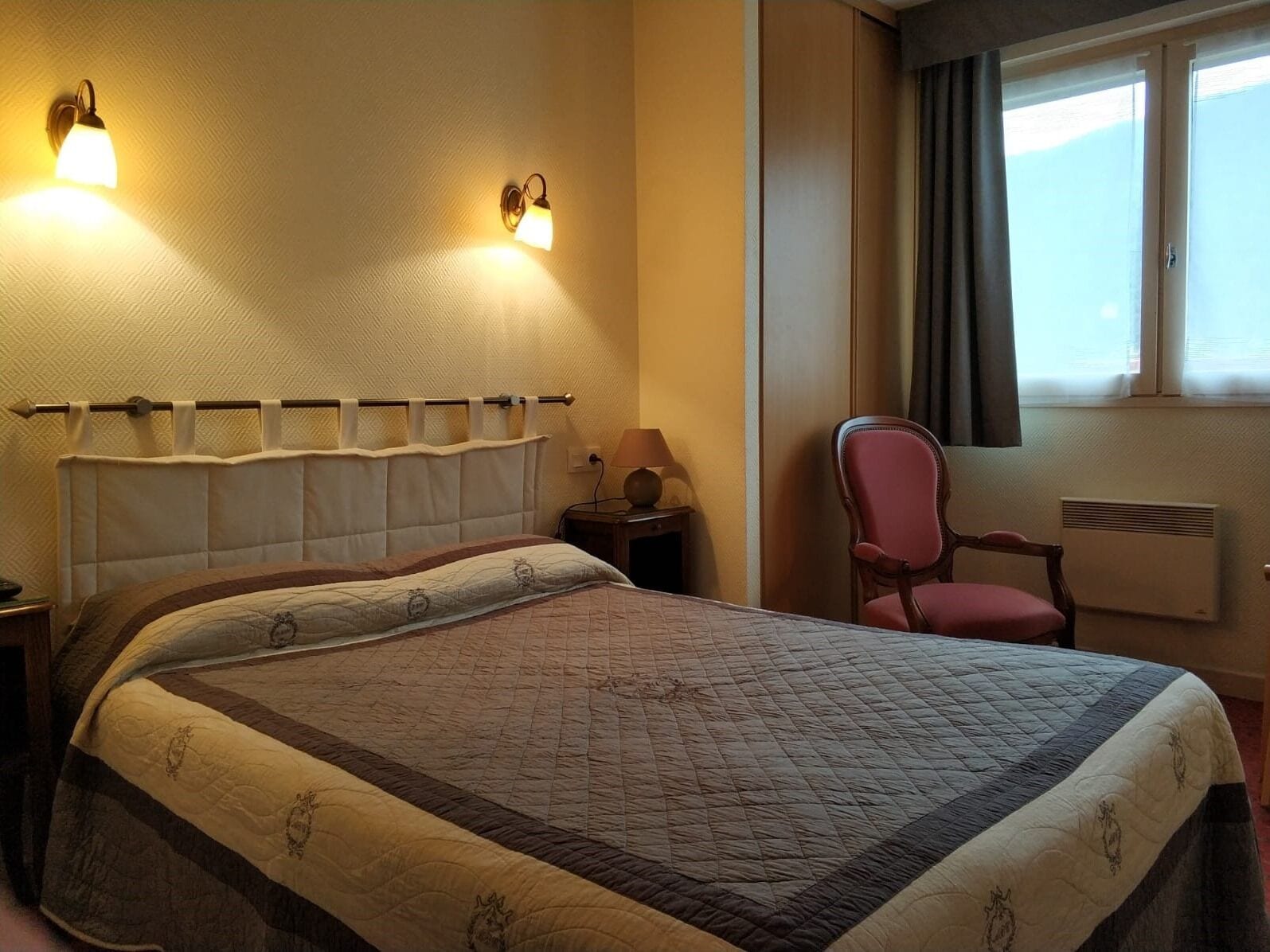 Quarto de hotel com uma cama, uma cadeira, uma mesa de cabeceira e duas luminárias na parede.