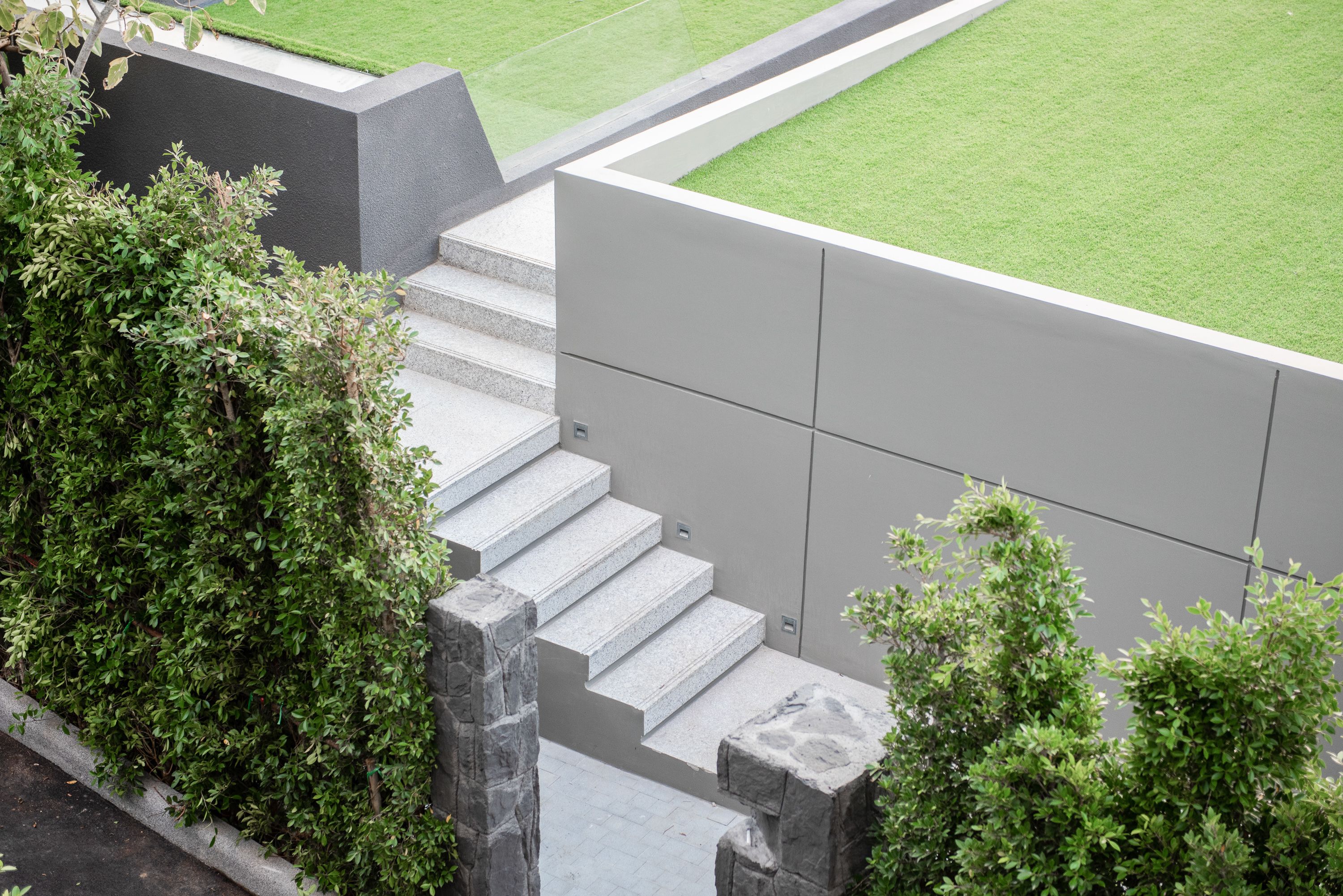 Uma escadaria moderna ao ar livre, com degraus de pedra que levam a um terraço plano, cercada por sebes verdes e um gramado bem cuidado.