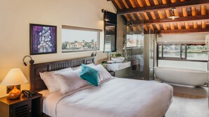 Quarto com teto de madeira, cama com roupa de cama branca, mesa de cabeceira com abajur, banheiro com banheira e janela com vista para prédios.