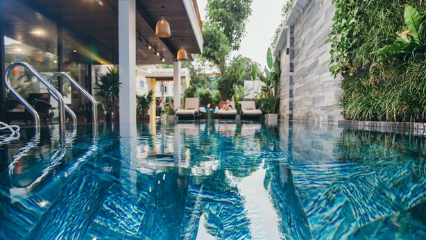 Uma piscina com água cristalina, cadeiras de descanso e um muro de pedra com vegetação.