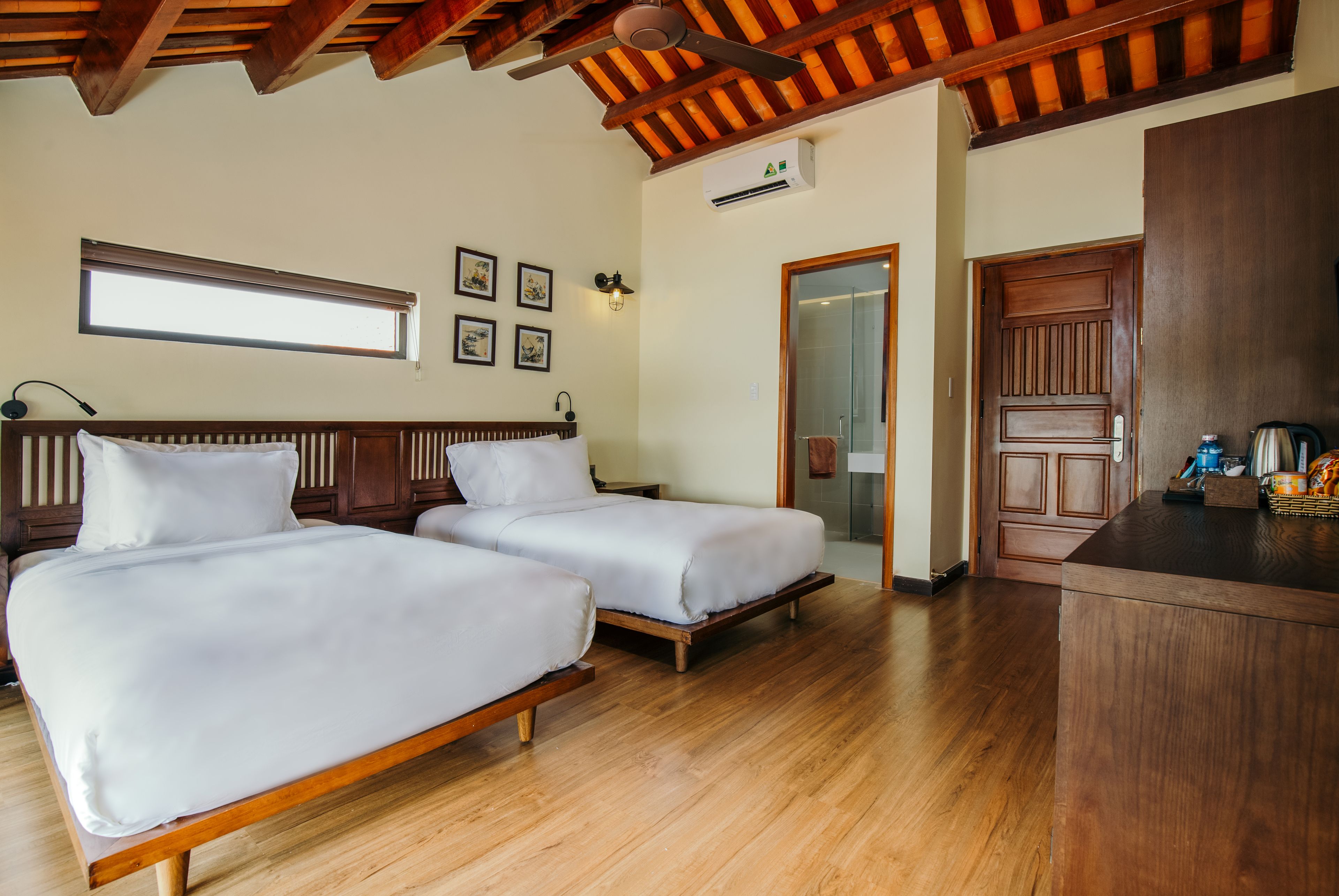 Quarto com duas camas, piso de madeira, ventilador de teto, uma janela e uma porta que dá para outro cômodo.