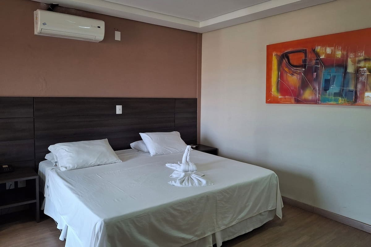 Quarto de hotel com cama, mesa de cabeceira, aparelho de ar condicionado e um quadro na parede.