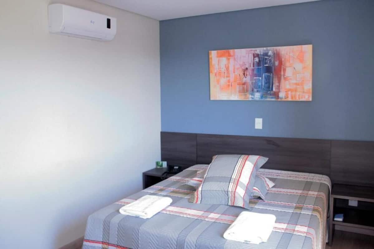 Quarto com cama, mesa de cabeceira, ar-condicionado na parede e um quadro.