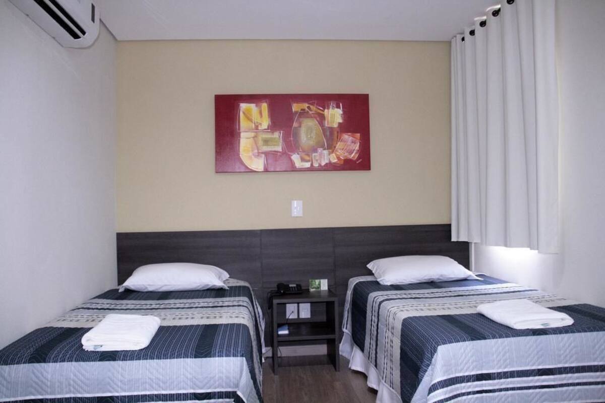 Quarto de hotel com duas camas, uma mesinha de cabeceira, uma obra de arte na parede e uma janela com cortinas.