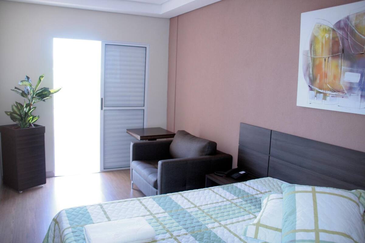 Quarto de hotel com cama, escrivaninha, cadeira, planta em vaso e porta de correr.