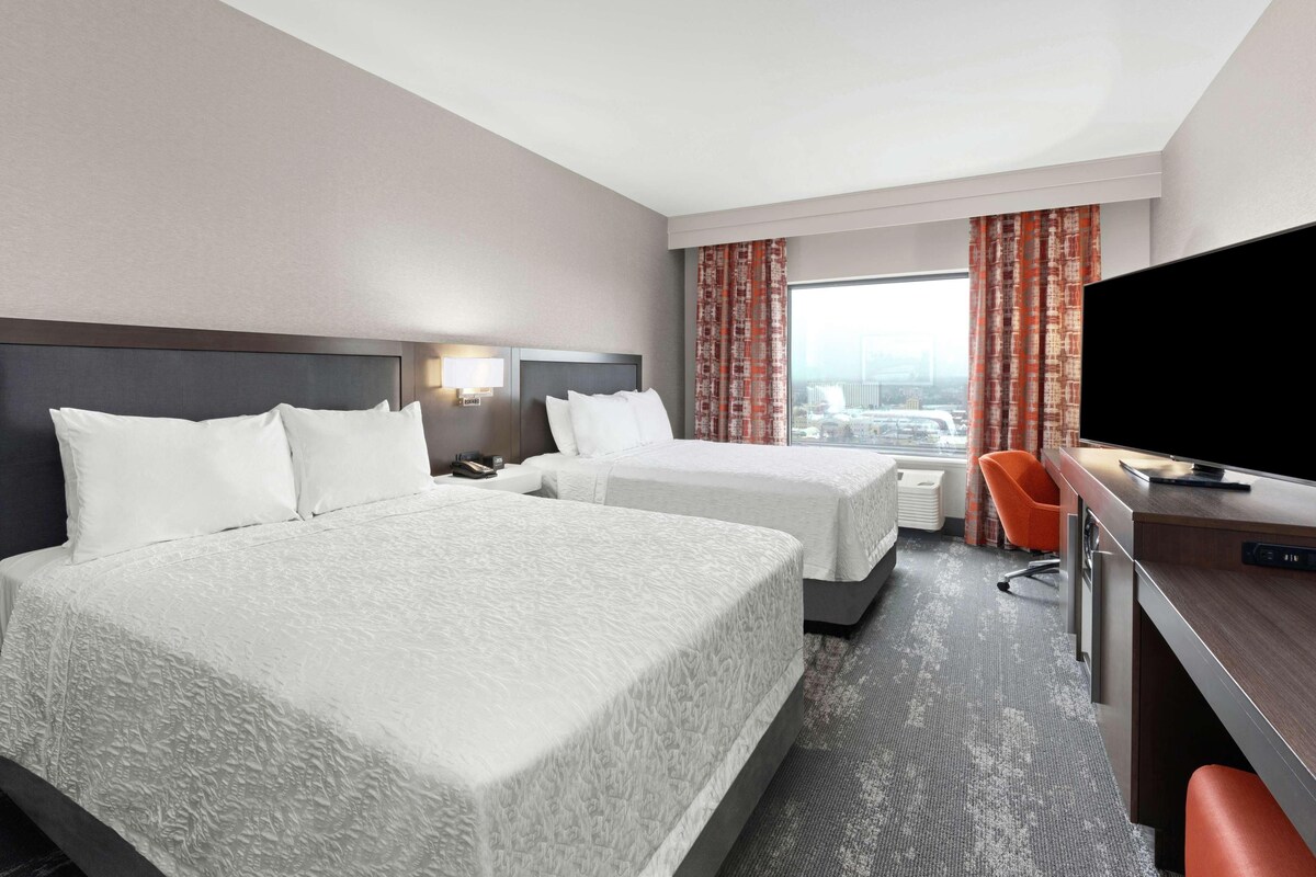 Quarto de hotel com duas camas, uma mesa de trabalho, televisão e vista para a cidade.