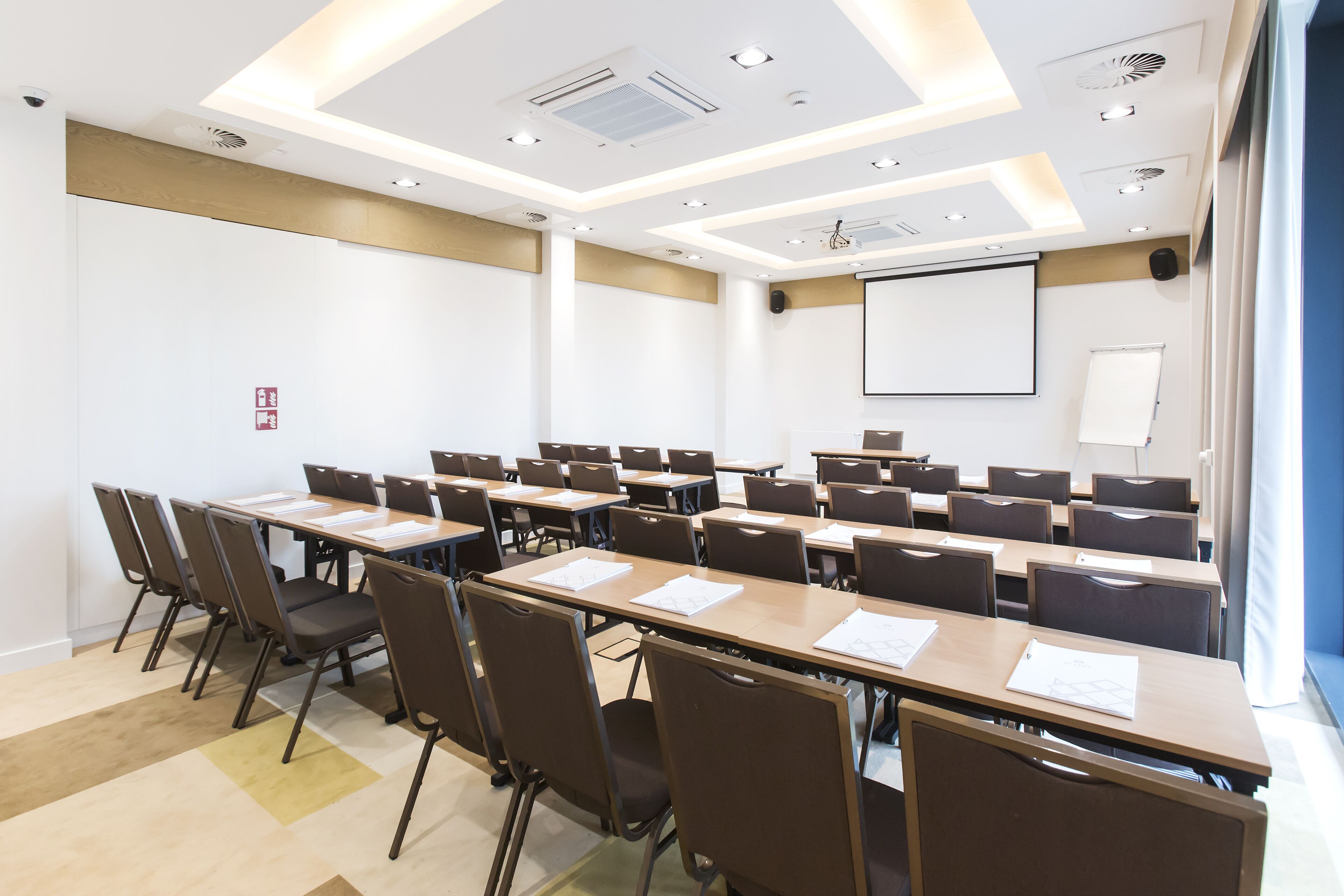 Uma sala de conferências com fileiras de mesas e cadeiras, uma tela de projetor e uma lousa.