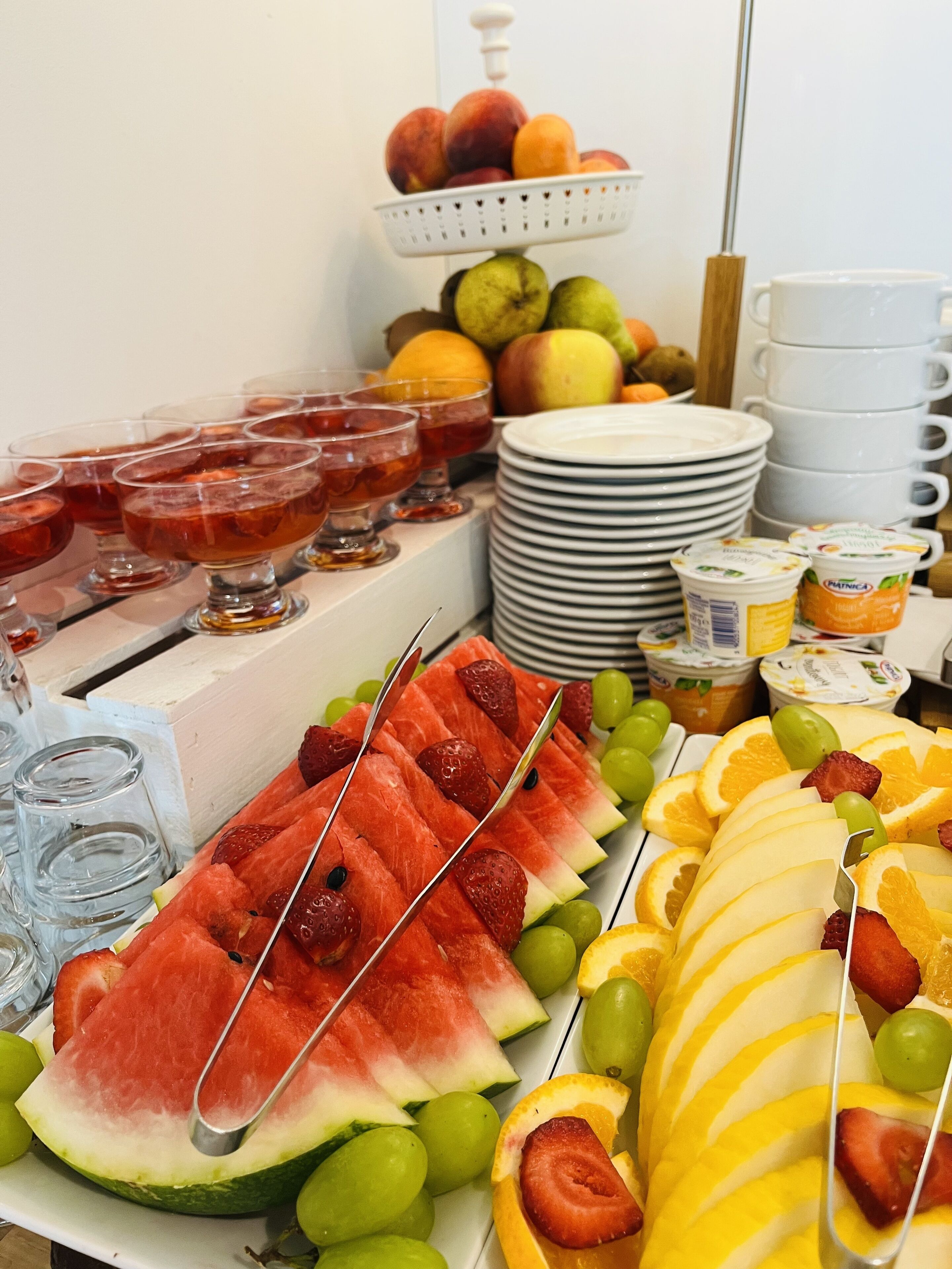 Buffet com diversas frutas, incluindo melancia, uvas e laranjas fatiadas, e vários recipientes de vidro com bebidas.