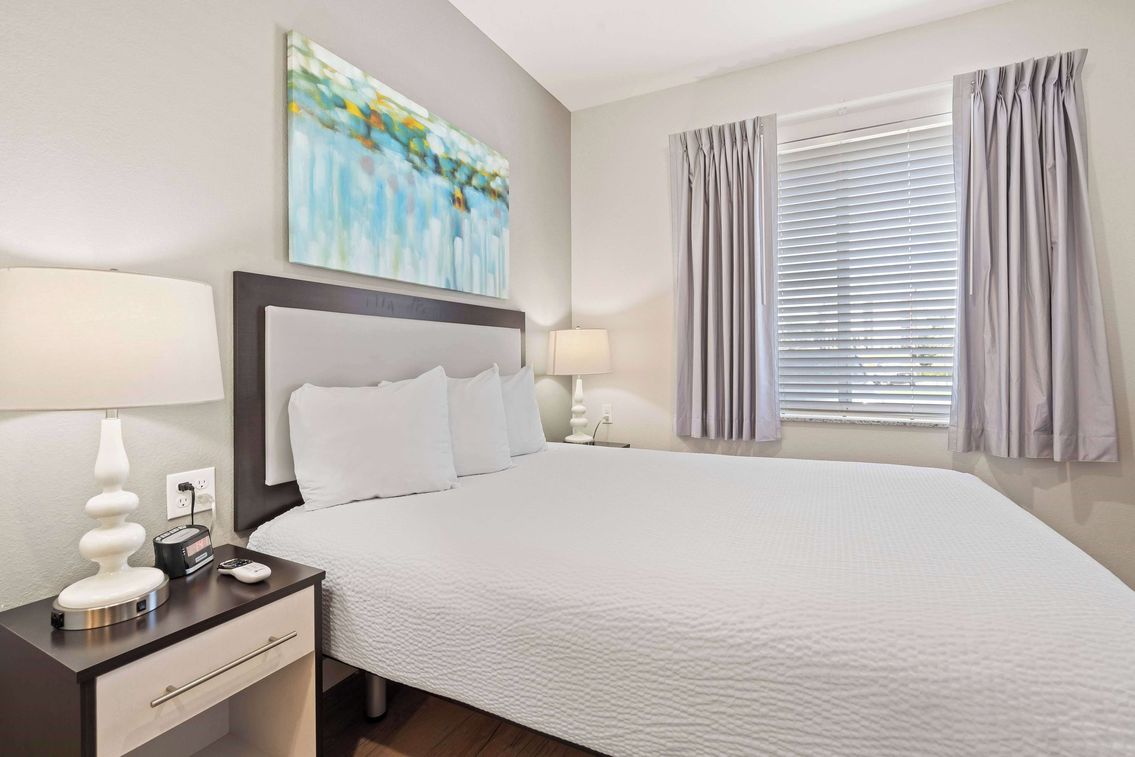 Quarto de hotel com uma cama grande, mesa de cabeceira com abajur, um quadro na parede e uma janela com cortinas.
