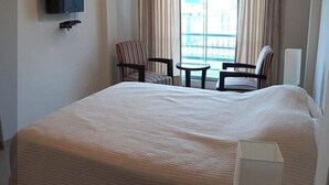 Quarto de hotel com uma cama grande, duas cadeiras, uma mesinha, televisão na parede e uma janela com cortinas.