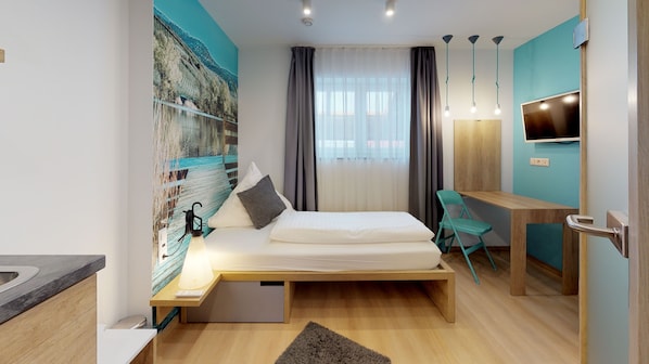 Quarto de hotel moderno com cama, escrivaninha, cadeira, televisão e uma janela grande com cortinas.