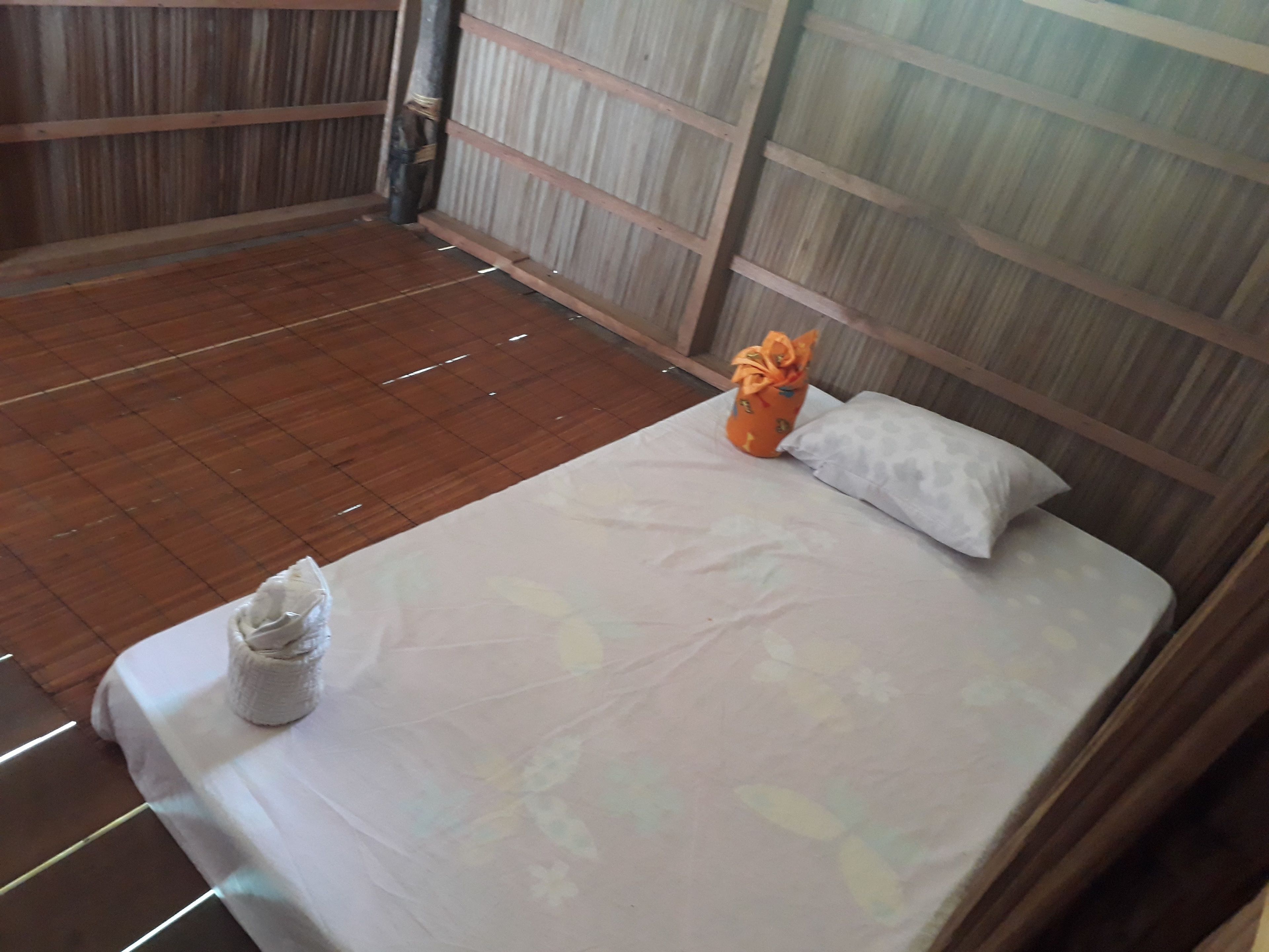 Uma cama de solteiro com lençol branco e padrão floral, travesseiro branco e um objeto decorativo laranja na cabeceira.