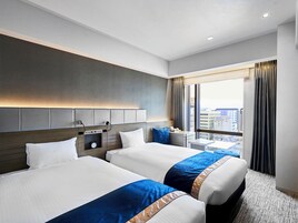 Quarto de hotel com duas camas, uma mesa de cabeceira, janela com cortinas e vista para a cidade.