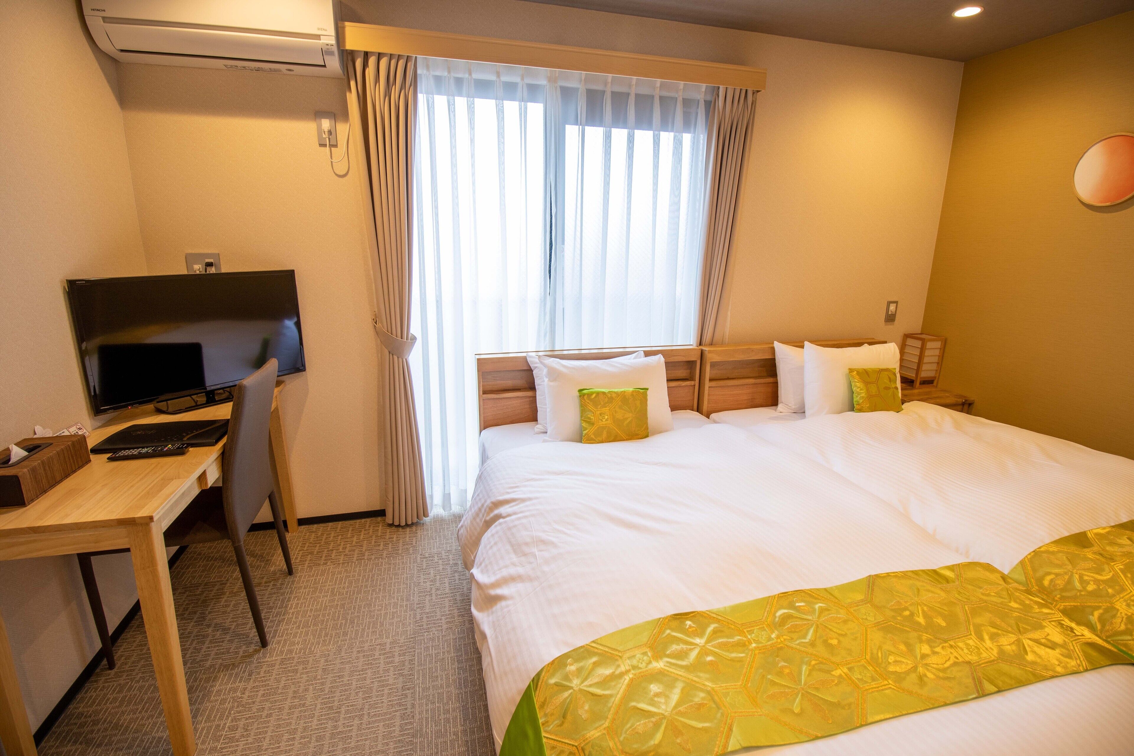 Quarto de hotel com cama, escrivaninha com computador, televisão e janela com cortinas.