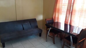 Um quarto com um sofá escuro, uma mesa de jantar com tampo de vidro e duas cadeiras de madeira.