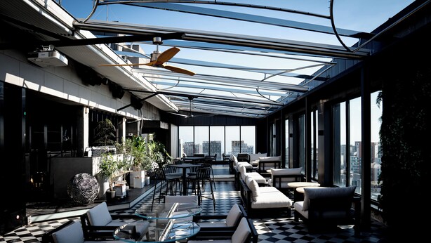 Um lounge no terraço com piso xadrez, mesas e cadeiras de vidro, e vista da cidade através de grandes janelas.