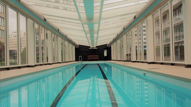 Piscina interior comprida e estreita, com água azul cristalina, rodeada por amplas janelas e um design moderno no teto.