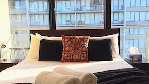 Quarto de hotel com cama, almofadas, cobertor e vista para um prédio pela janela.